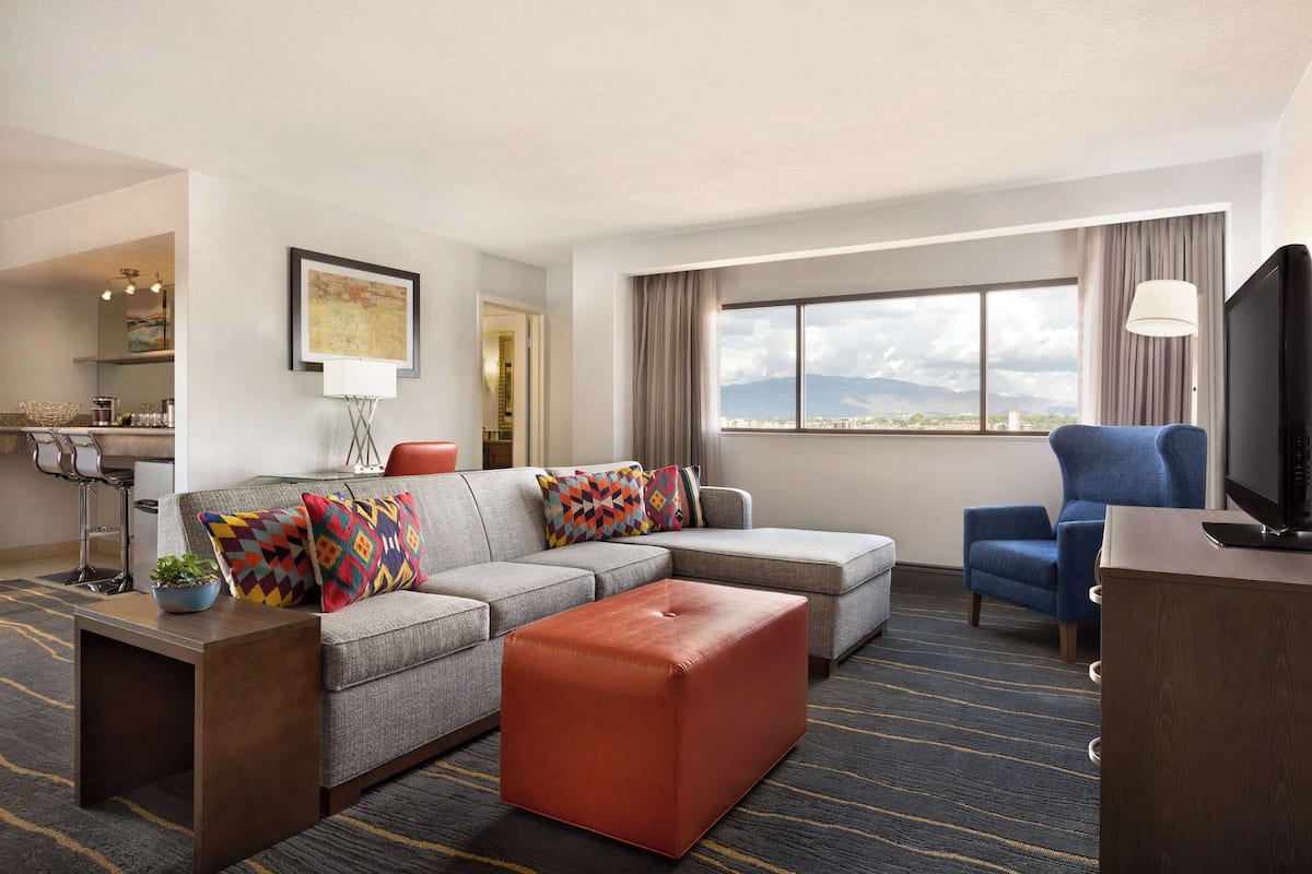 Quarto de hotel com sofá seccional, poltrona azul, televisão e janela grande com vista para a cidade.