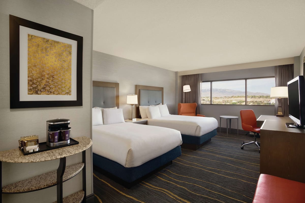 Quarto de hotel com duas camas, uma escrivaninha com cadeira, televisão e janela com vista para o deserto.