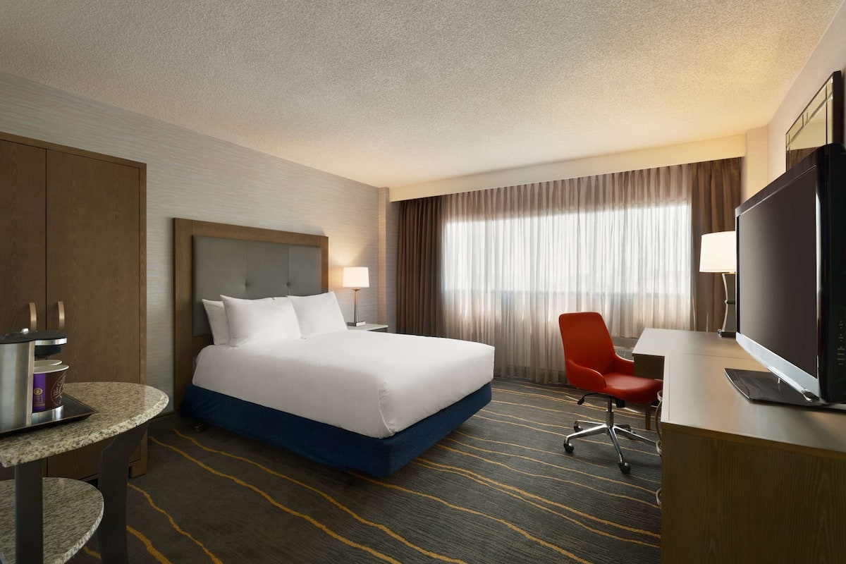 Quarto de hotel com uma cama grande, uma escrivaninha com computador, uma TV e uma cadeira.