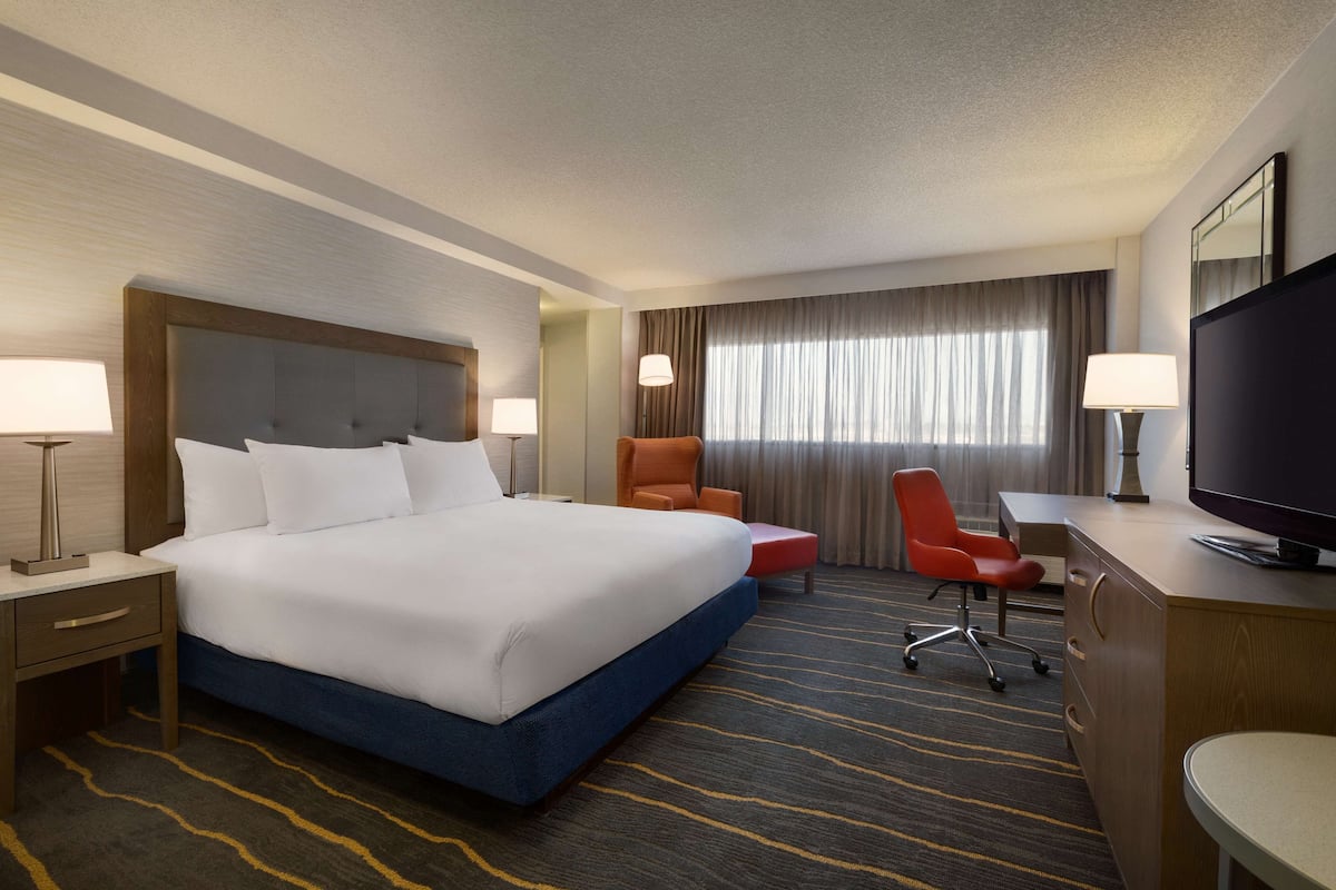 Quarto de hotel com uma cama grande, duas cadeiras, uma mesa de trabalho e uma televisão, além de uma janela com cortinas.