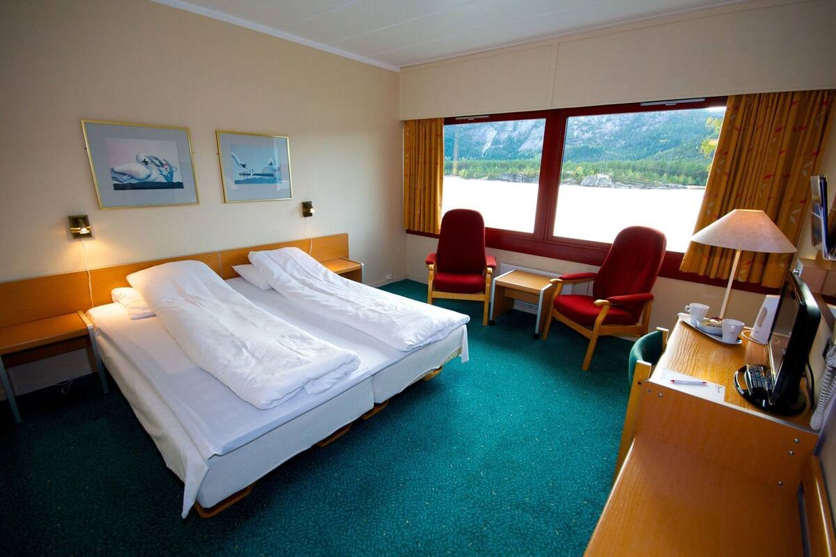 Quarto de hotel com cama, duas cadeiras, escrivaninha, abajur, telefone e janela com vista para as montanhas.