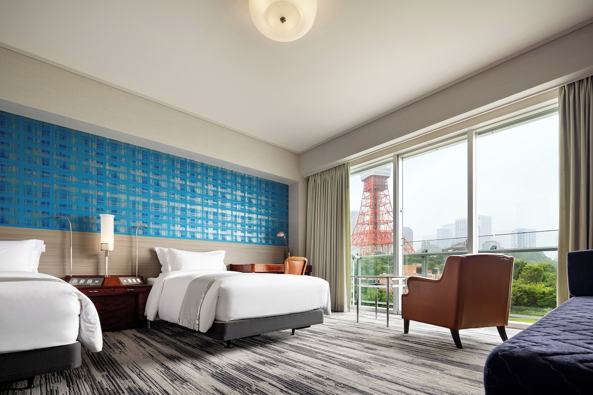Quarto de hotel com uma cama grande, uma cama menor, uma cadeira, uma escrivaninha e uma janela ampla com vista para a cidade.