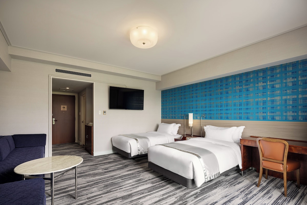 Quarto de hotel com duas camas, uma escrivaninha, uma cadeira, uma mesinha e um grande painel na parede.