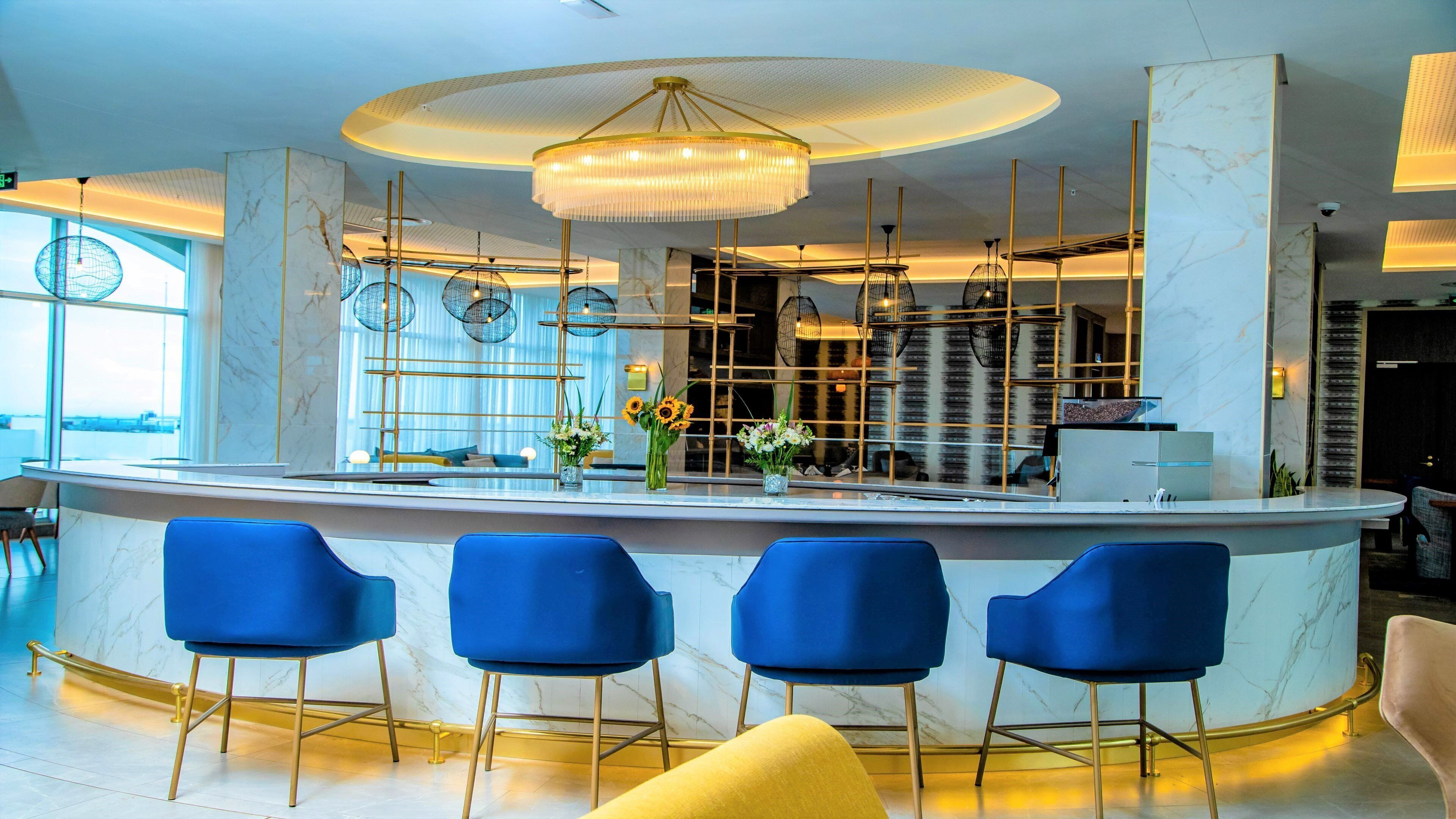 Uma área de bar moderna com uma ilha central, banquetas azuis e um grande candelabro.