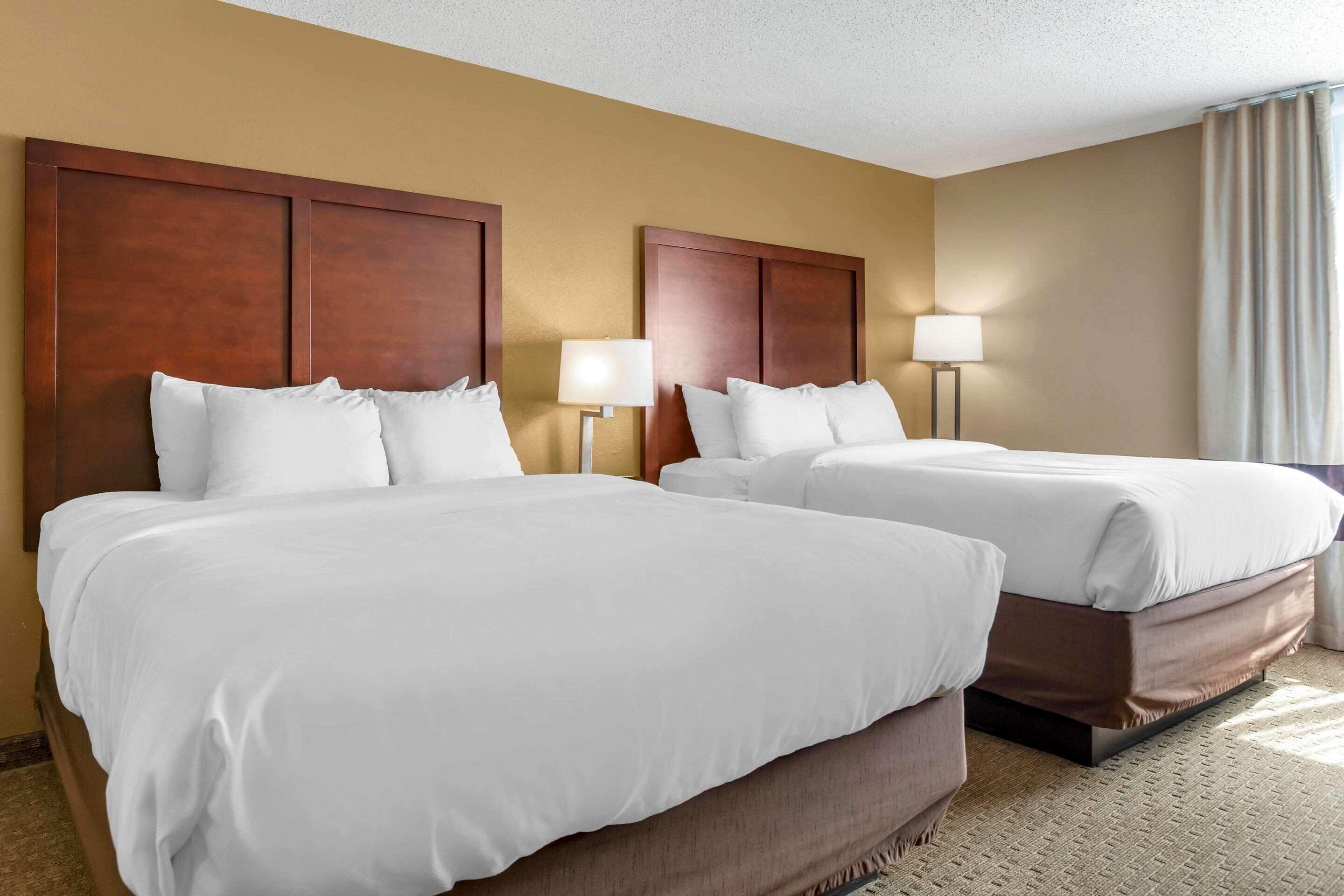Quarto de hotel com duas camas, cada uma com roupa de cama branca e cabeceiras de madeira. Possui duas luminárias de cabeceira e uma janela com cortinas.