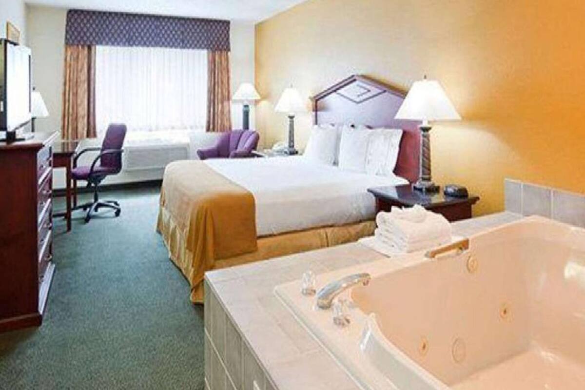 Quarto de hotel com uma cama grande, área de estar separada com uma poltrona, escrivaninha, luminária de mesa, mesa de cabeceira com abajur e banheira.
