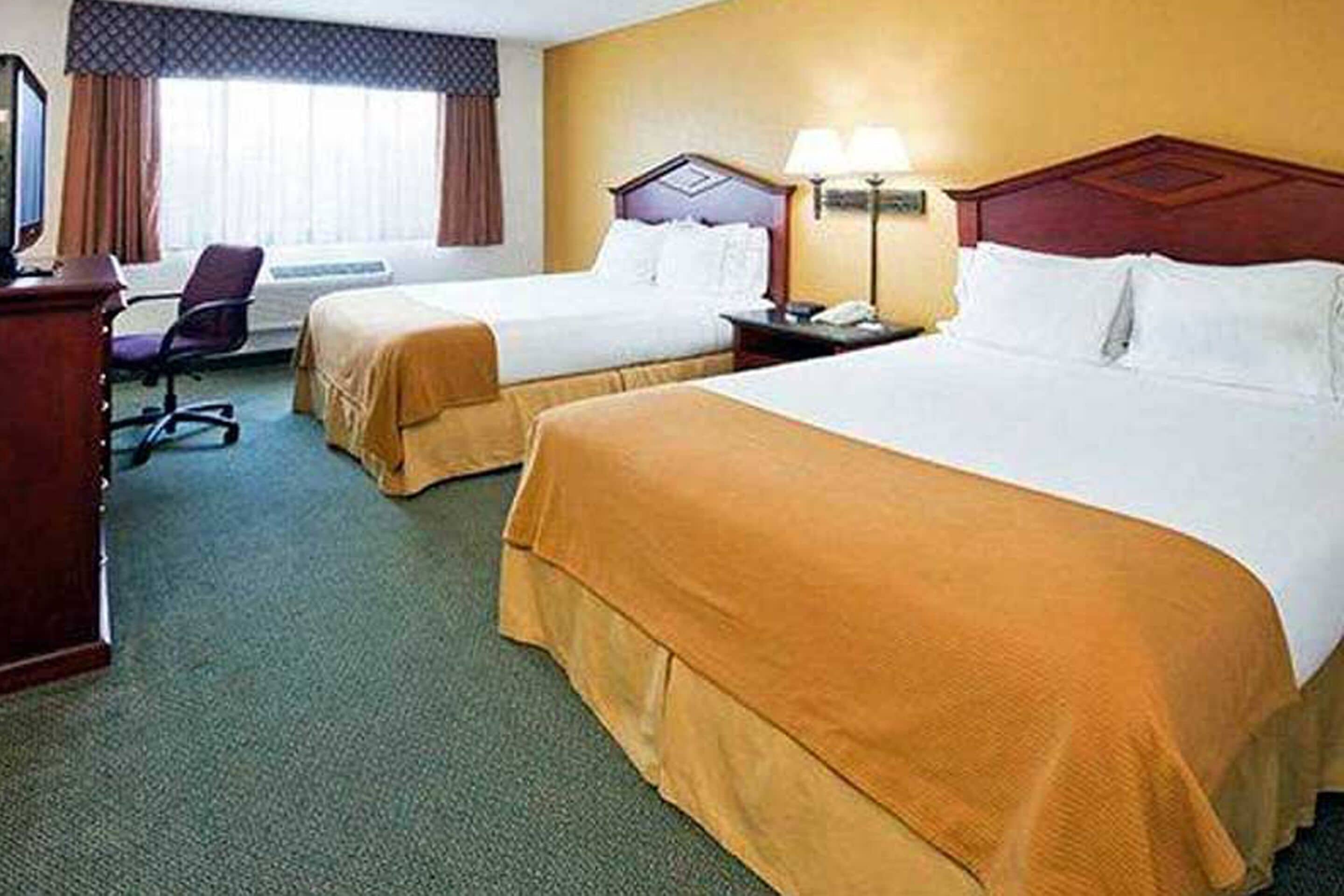 Quarto de hotel com duas camas, uma escrivaninha, uma cadeira e uma janela com cortinas.
