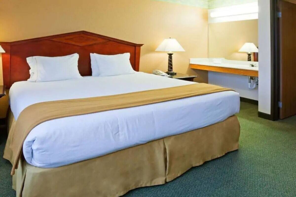 Quarto de hotel com uma cama grande, duas luminárias laterais, um painel de cabeceira de madeira, um tapete verde e um banheiro com pia e espelho.
