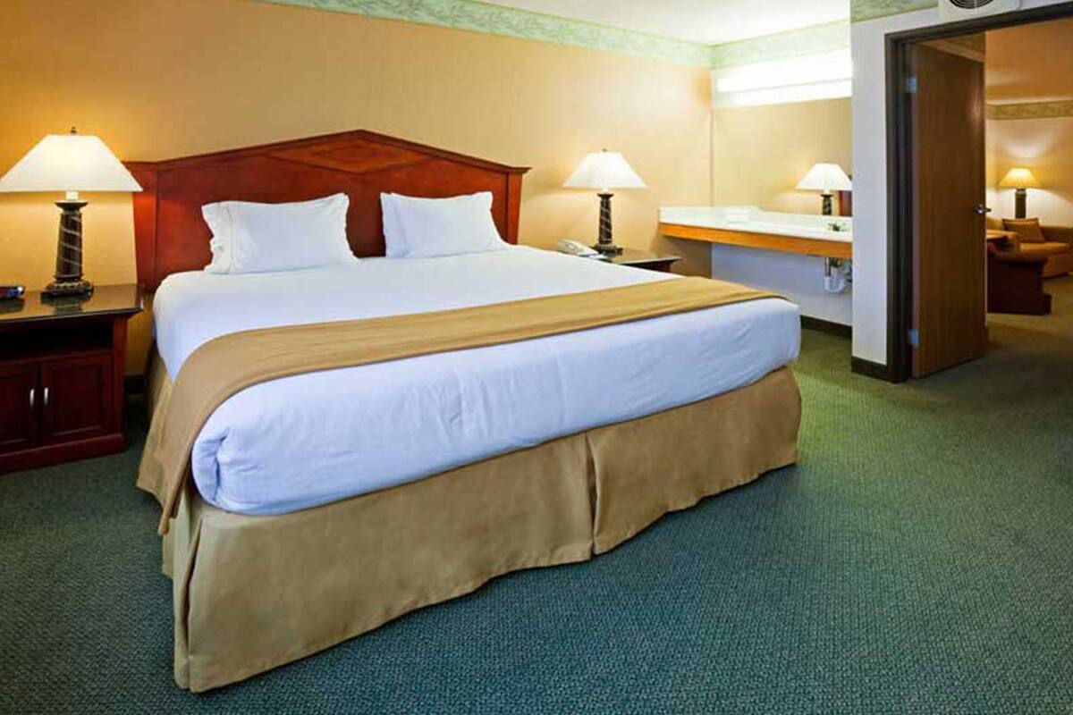 Quarto de hotel com uma cama grande, duas mesas de cabeceira com luminárias, um painel de cama de madeira, uma escrivaninha com cadeira e um tapete verde.
