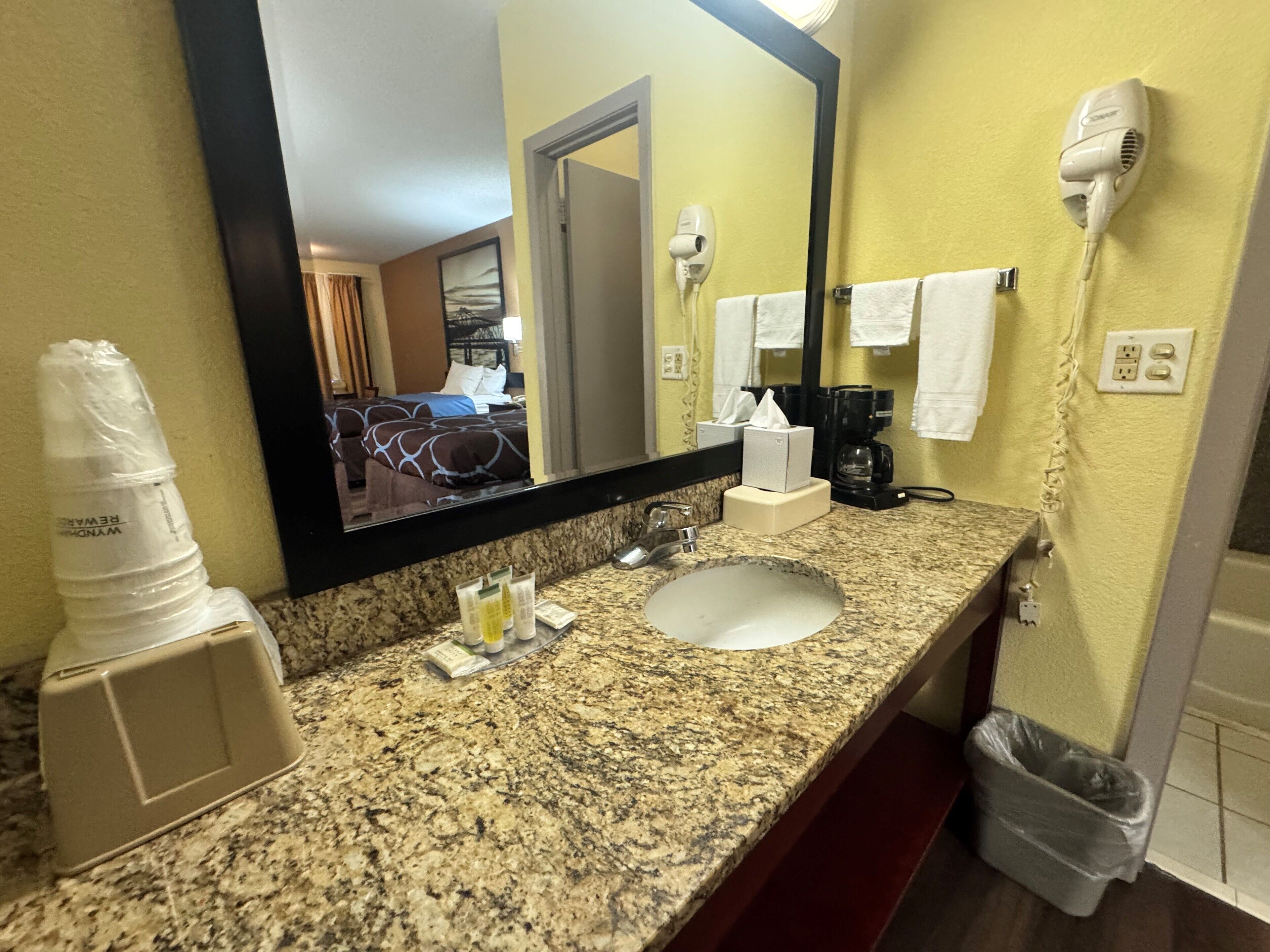 Casa de banho de hotel com bancada de granito, pia, espelho, secador de cabelo, porta-toalhas e um cesto de lixo.