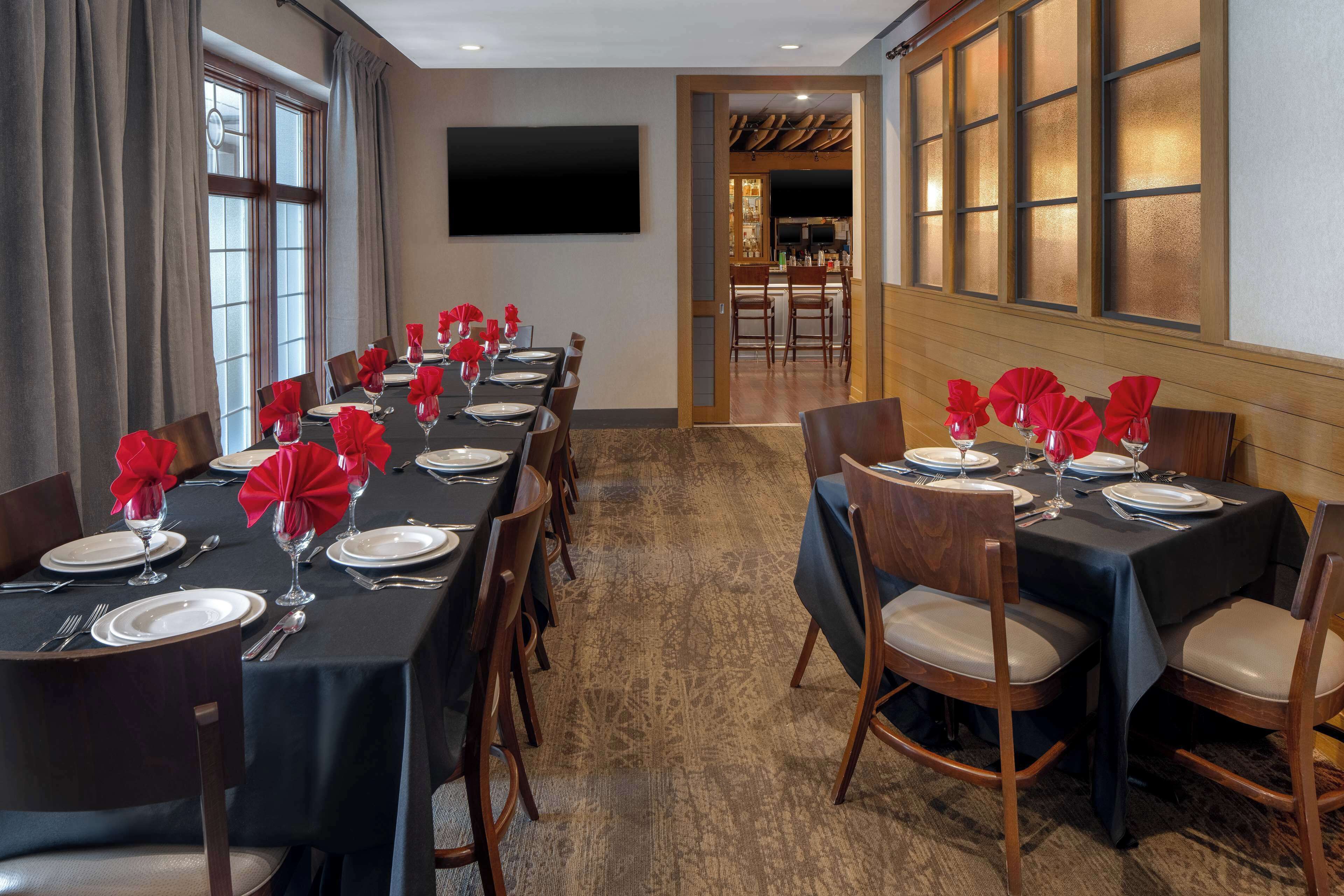 Sala de conferências com mesas compridas preparadas para um evento formal, uma grande TV de tela plana na parede e vista para a cozinha através de uma porta de vidro.