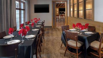 Sala de conferências com mesas compridas preparadas para um evento formal, uma grande TV de tela plana na parede e vista para a cozinha através de uma porta de vidro.