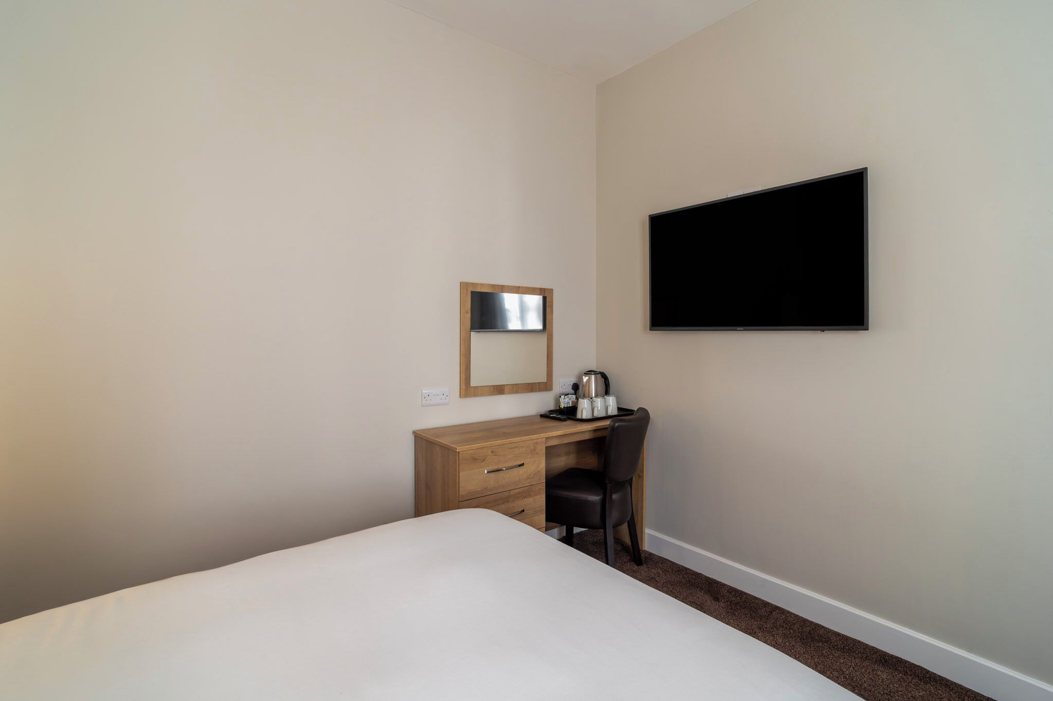Quarto de hotel com uma cama, uma escrivaninha com cadeira, uma TV de tela plana na parede e uma mesinha com cafeteira.