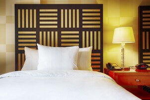 Quarto de hotel com cama, travesseiros, mesa de cabeceira com abajur, telefone e parede com padrão geométrico.