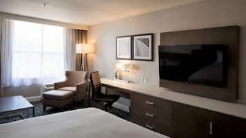 Quarto de hotel com uma cama grande, uma escrivaninha, uma cadeira, uma televisão e uma janela com cortinas.