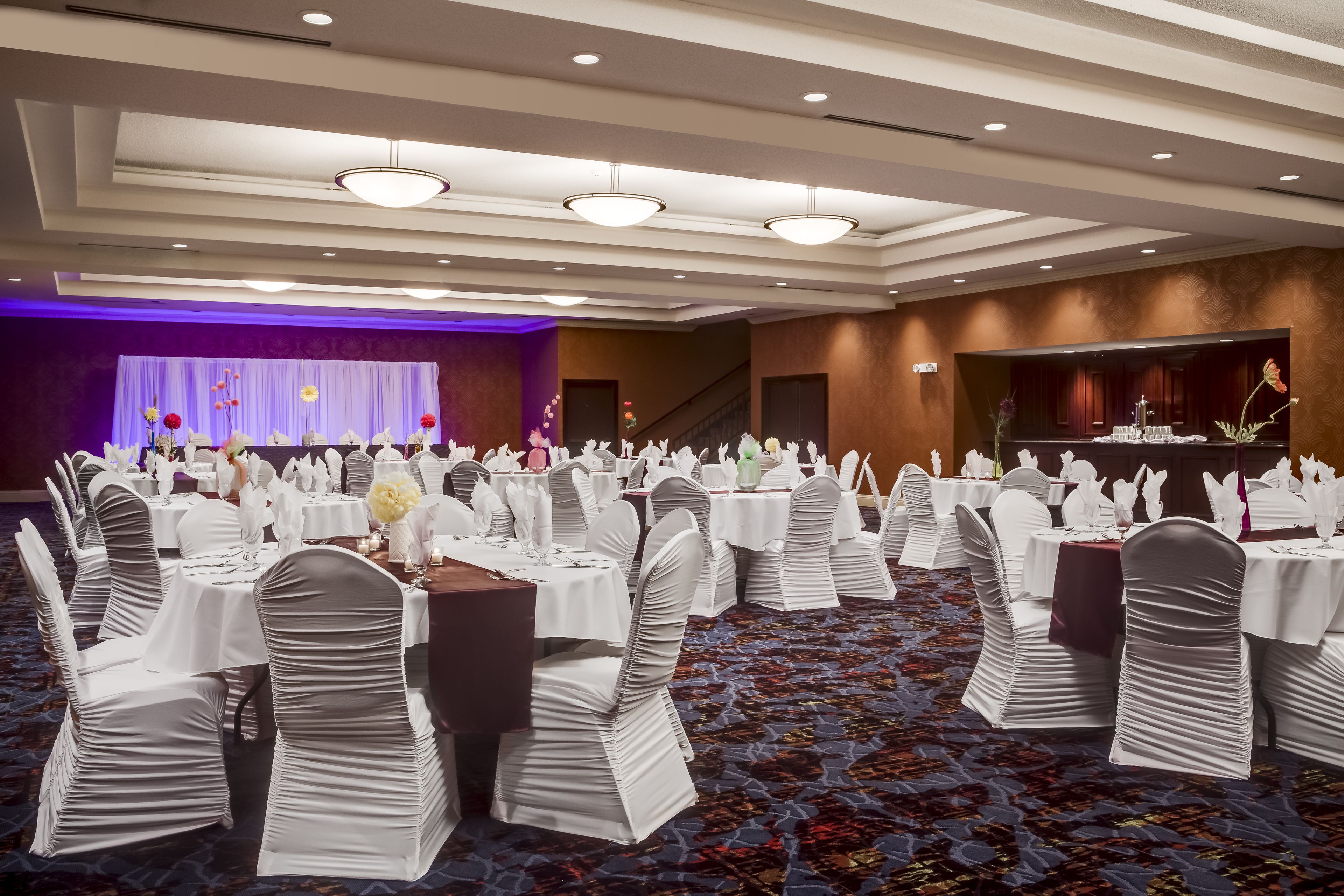 Uma sala de banquetes com mesas redondas, toalhas brancas e cadeiras adornadas com fitas brancas.