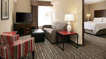 Quarto de hotel com sofá, poltrona, televisão, mesa de cabeceira, luminária e cama.