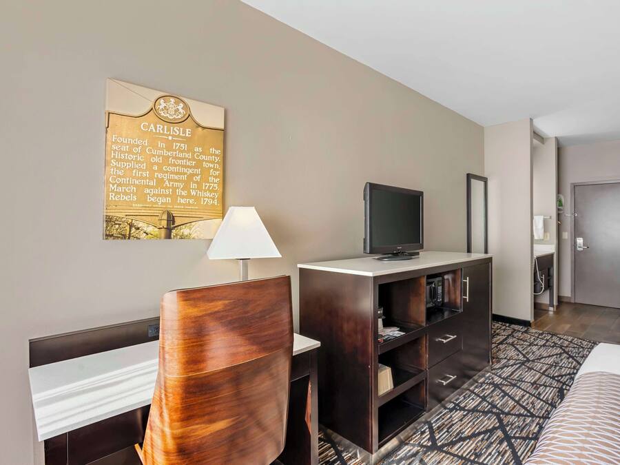 Quarto de hotel com uma cadeira de madeira, uma escrivaninha, uma televisão, uma estante e uma placa histórica emoldurada na parede.