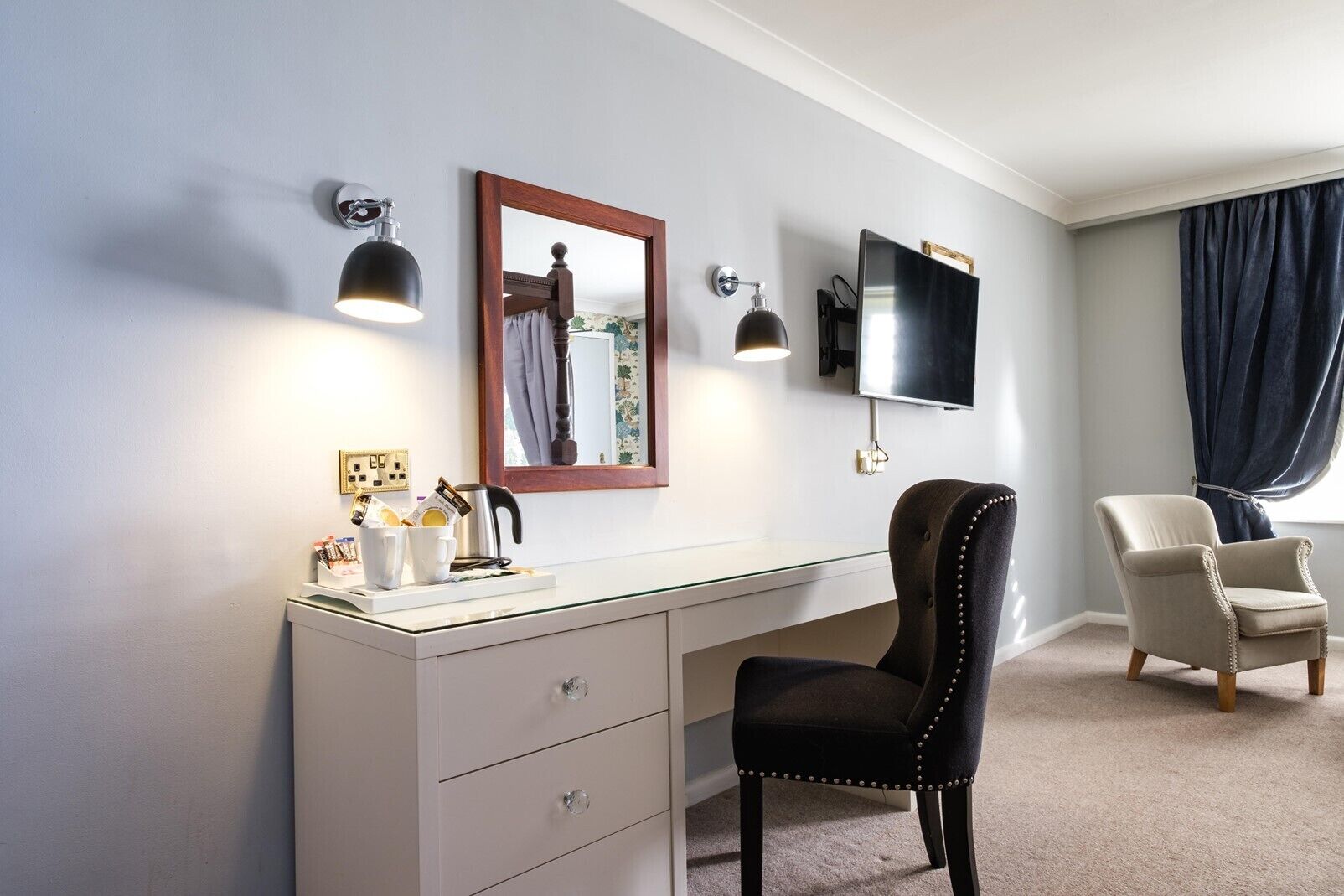 Quarto de hotel com uma escrivaninha branca, uma cadeira preta, uma televisão e um espelho na parede.