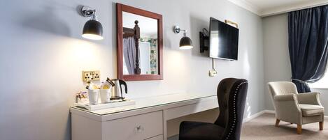 Quarto de hotel com uma escrivaninha branca, uma cadeira preta, uma televisão e um espelho na parede.