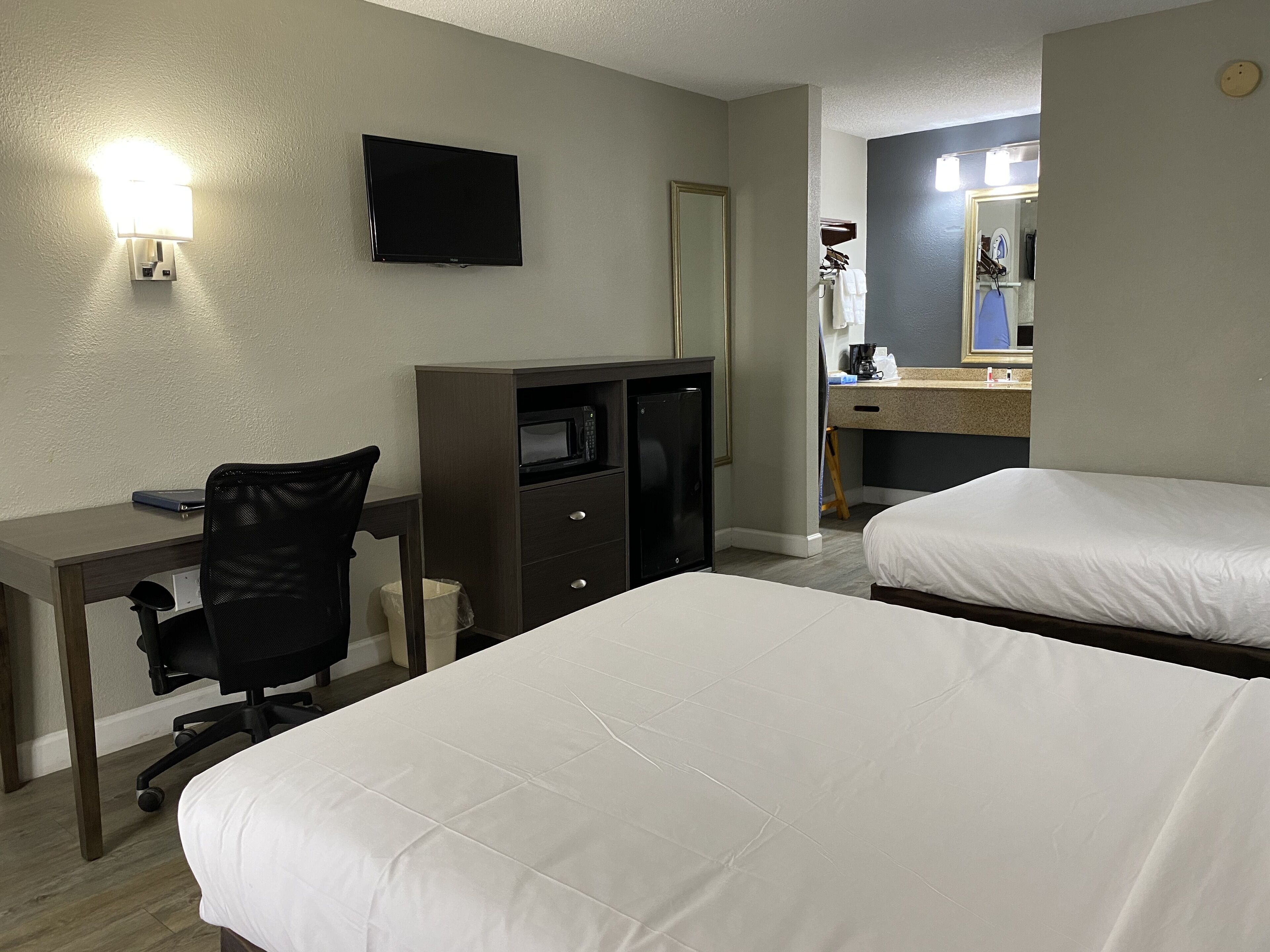 Quarto de hotel com duas camas, uma mesa com cadeira, TV fixada na parede e pequena área de cozinha.