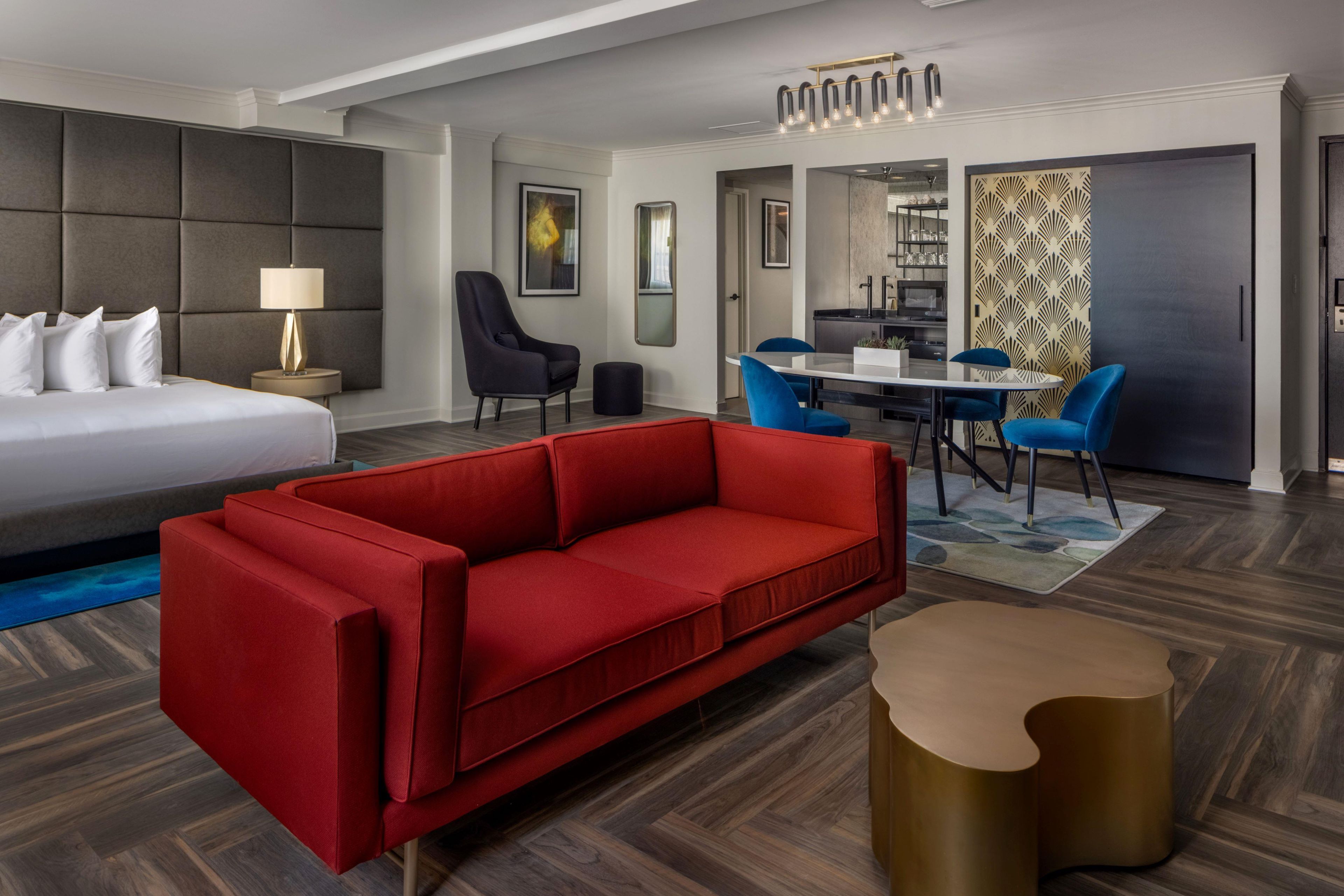 Quarto de hotel com cama, sofá vermelho, mesa redonda pequena, cadeira, abajur e área de refeições com mesa e cadeiras.