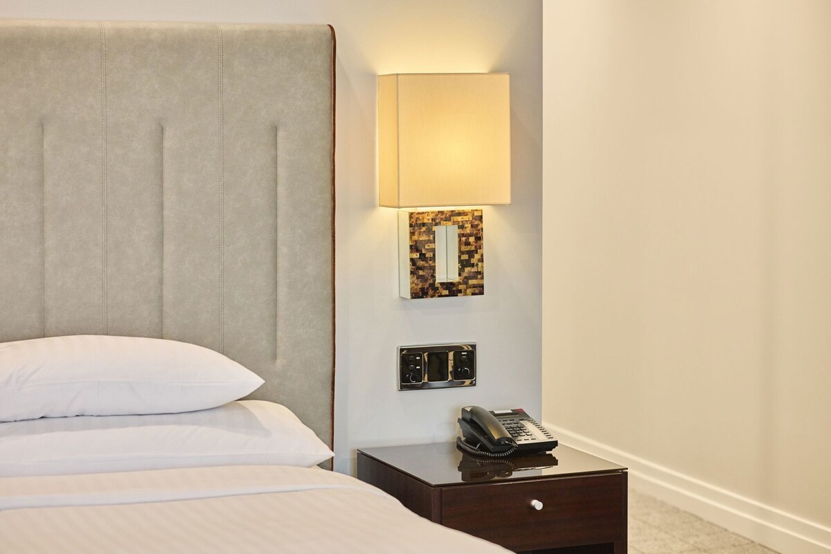 Cama bem arrumada com cabeceira, abajur, telefone e luminária na parede.