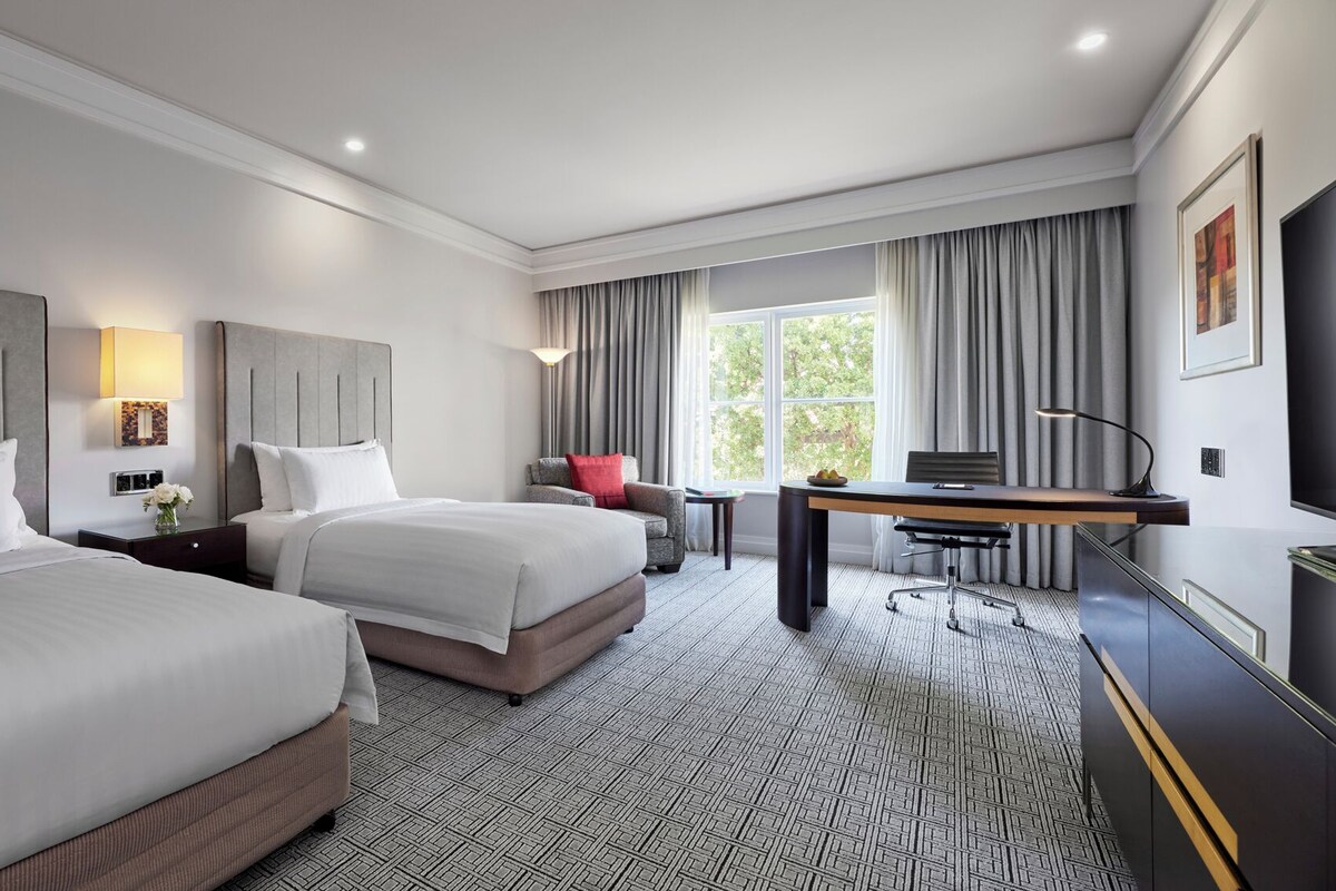 Quarto de hotel com duas camas, uma escrivaninha, uma cadeira, uma televisão e uma janela com cortinas.