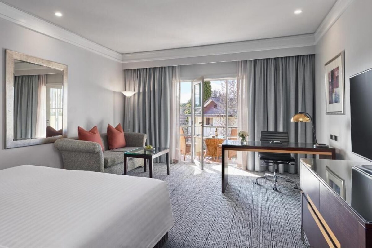Quarto de hotel com uma cama grande, uma mesa, uma cadeira, televisão e varanda com cortinas.