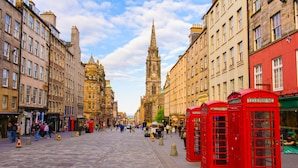 Uma rua de paralelepípedos ladeada por edifícios históricos, com uma torre imponente e cabines telefônicas vermelhas.