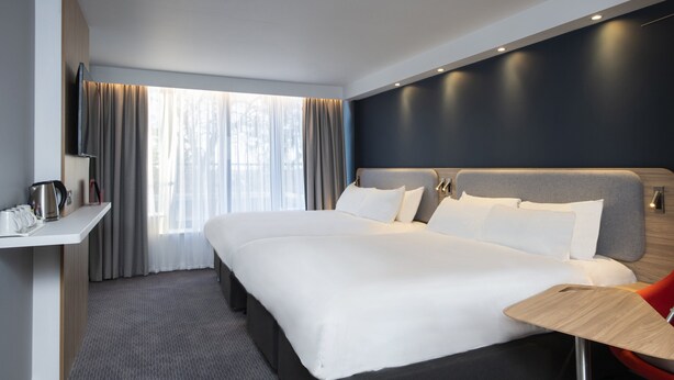 Quarto de hotel moderno com uma cama grande, uma mesa de cabeceira, uma escrivaninha e vista para o exterior pela janela.