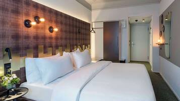 Quarto de hotel com uma cama grande, uma mesa de cabeceira com abajur, luminária na parede e uma porta que dá para outro quarto.