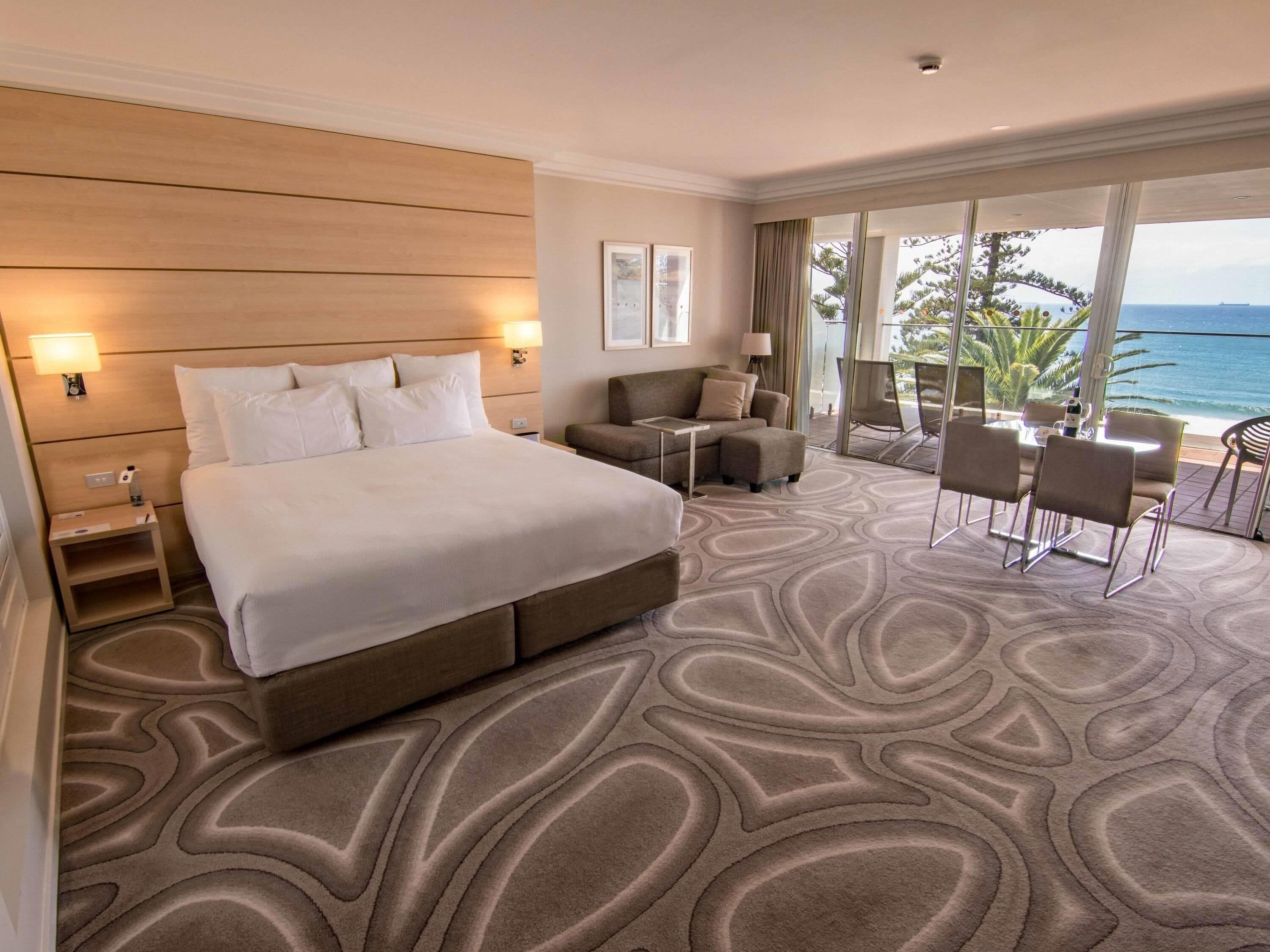 Quarto de hotel com uma cama grande, um sofá, uma mesinha e vista para o oceano.