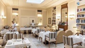 Uma sala de jantar com várias mesas e cadeiras, um chef preparando comida e uma estante repleta de livros.