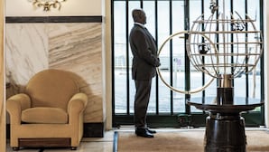 Um homem está em um saguão com um candelabro, uma escultura de globo terrestre e uma grande janela em arco.
