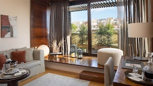 Uma sala de estar moderna com um sofá, uma mesa de centro, uma mesinha de apoio, uma luminária e vista para a cidade por grandes janelas.
