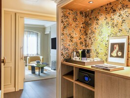 Quarto de hotel com secretária de madeira, cafeteira, chaleira, quadro decorativo e estante com um livro intitulado
