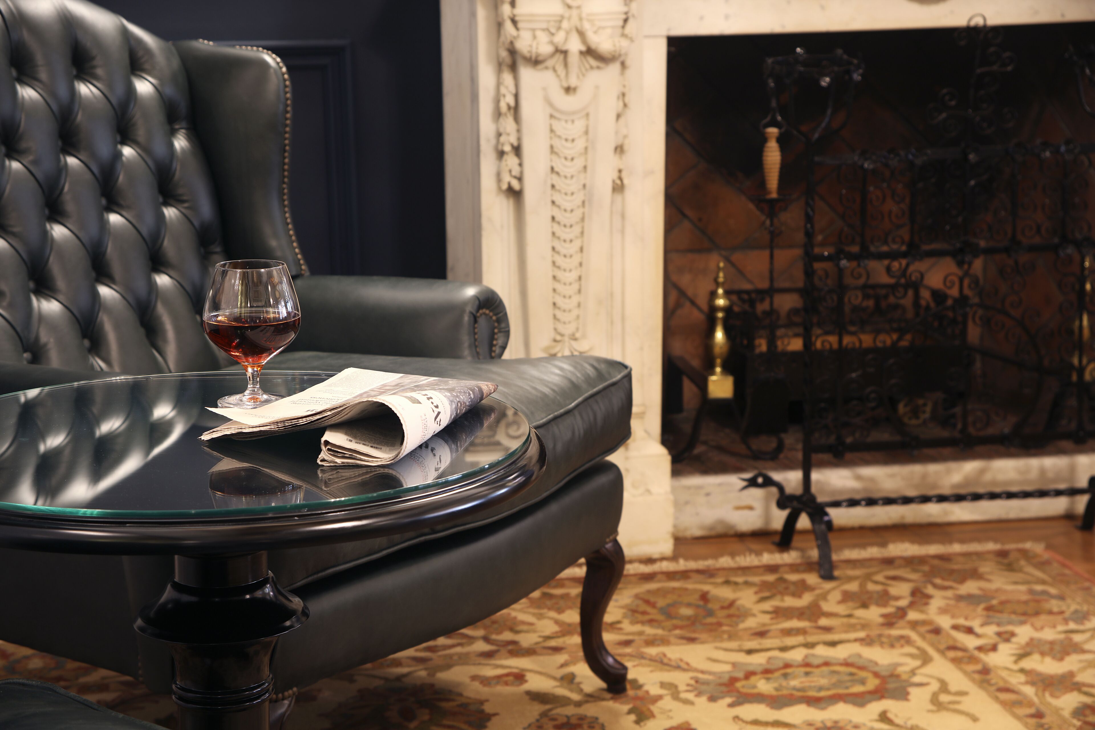 Uma poltrona de couro, uma mesa de vidro com um jornal e uma lareira com portão decorativo de ferro.