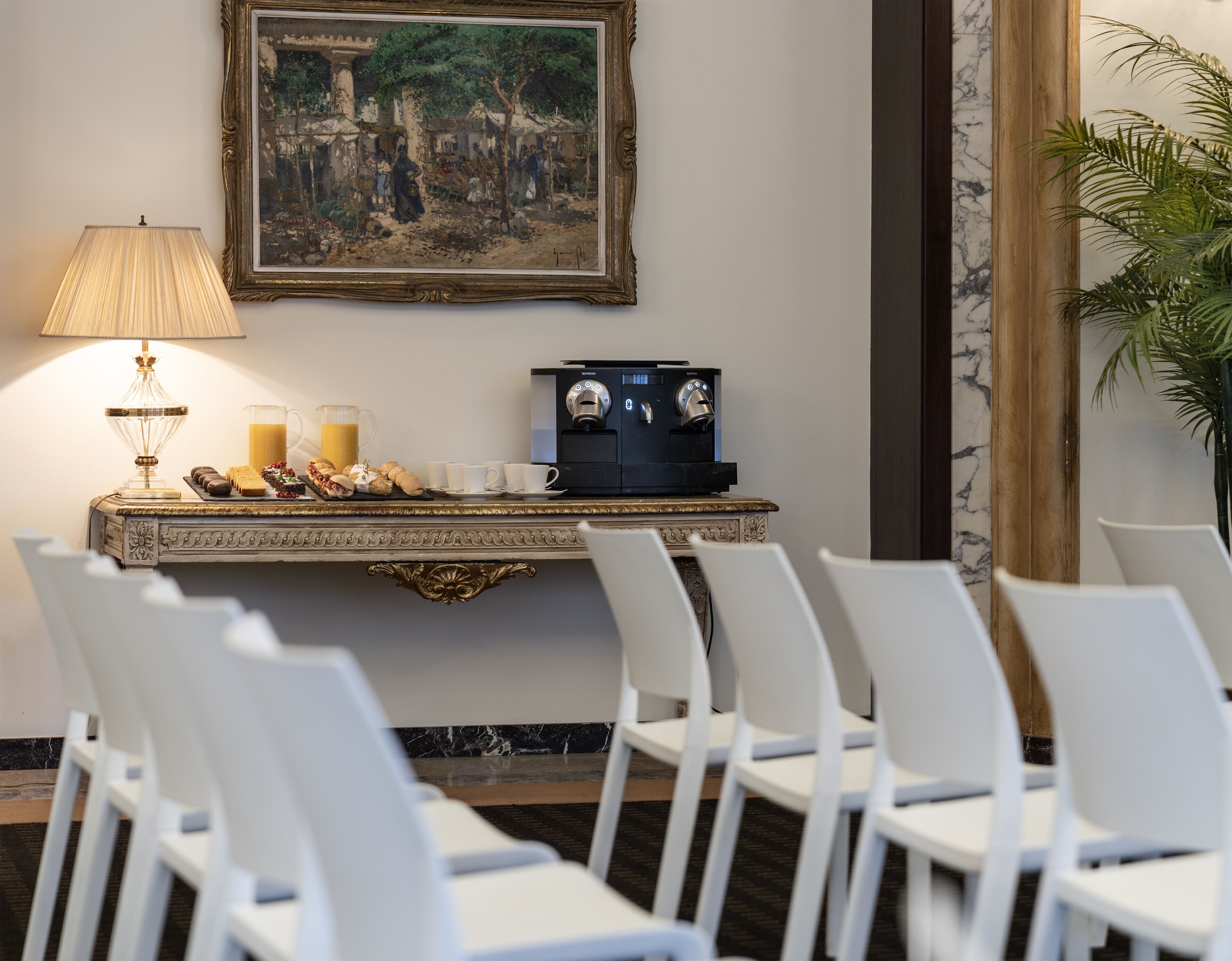 Uma sala de jantar com cadeiras brancas, uma máquina de café, um abajur e um quadro na parede.