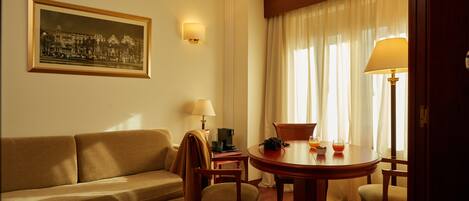 Quarto de hotel com sofá, mesa de jantar com cadeiras, luminária, pintura na parede e janela com cortinas.
