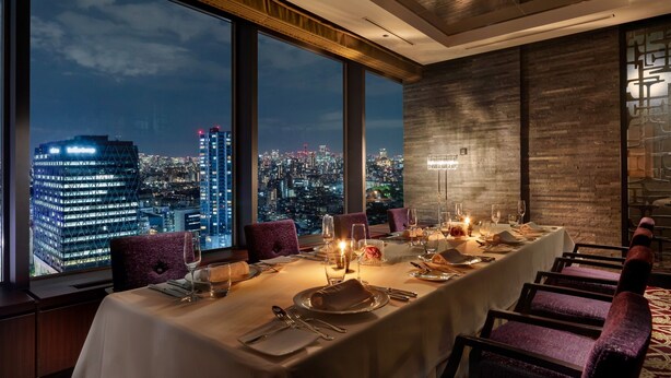 Uma sala de jantar com vista para a cidade, grandes janelas e uma mesa comprida posta para refeição.