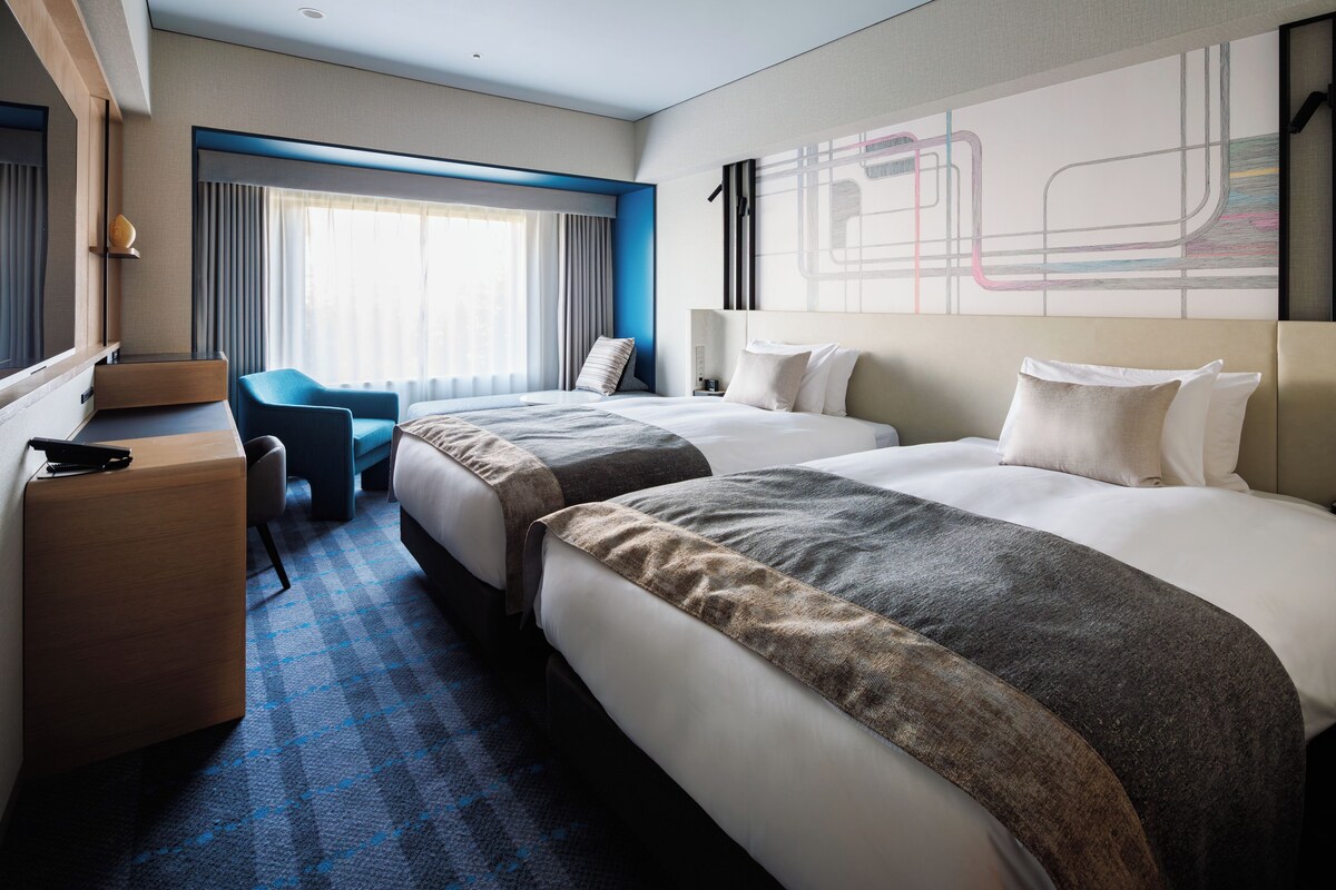 Quarto de hotel com duas camas, uma escrivaninha, uma cadeira e uma janela grande com cortinas.