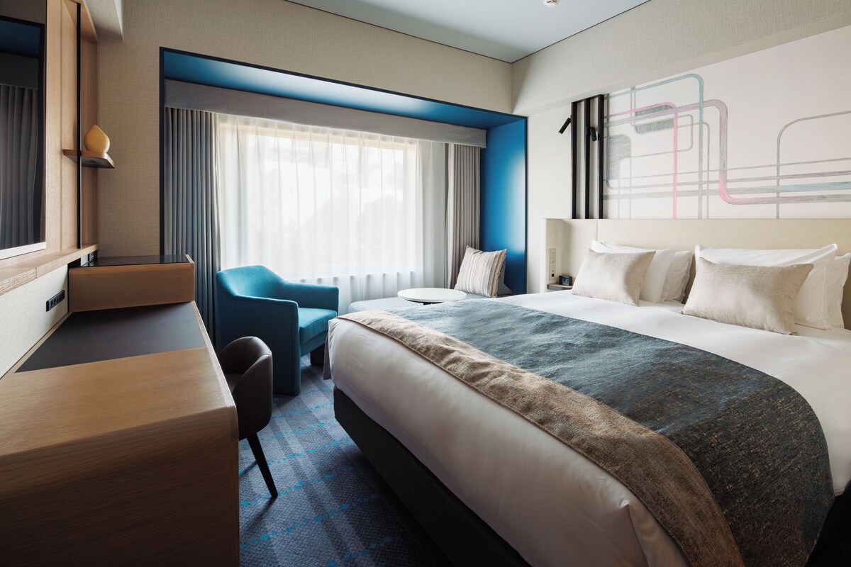 Quarto de hotel moderno com uma cama grande, uma escrivaninha, uma cadeira e uma janela com cortinas.