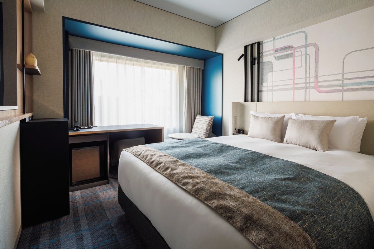 Quarto de hotel com cama, escrivaninha, cadeira e uma grande janela com cortinas.