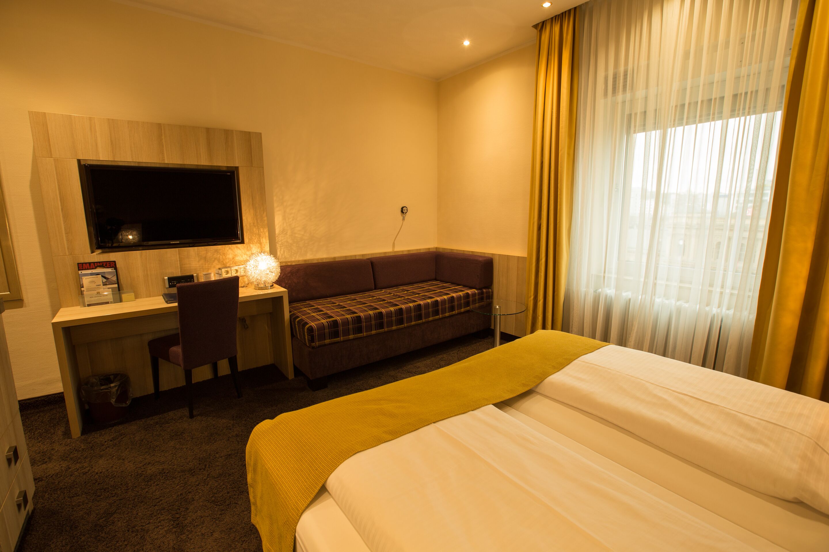Quarto de hotel com cama, escrivaninha com computador, cadeira, sofá e televisão na parede.