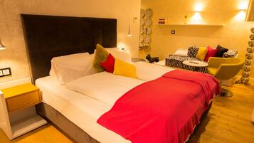 Quarto de hotel com uma cama grande, uma mesinha de cabeceira, uma cadeira e uma pequena mesa com abajur.