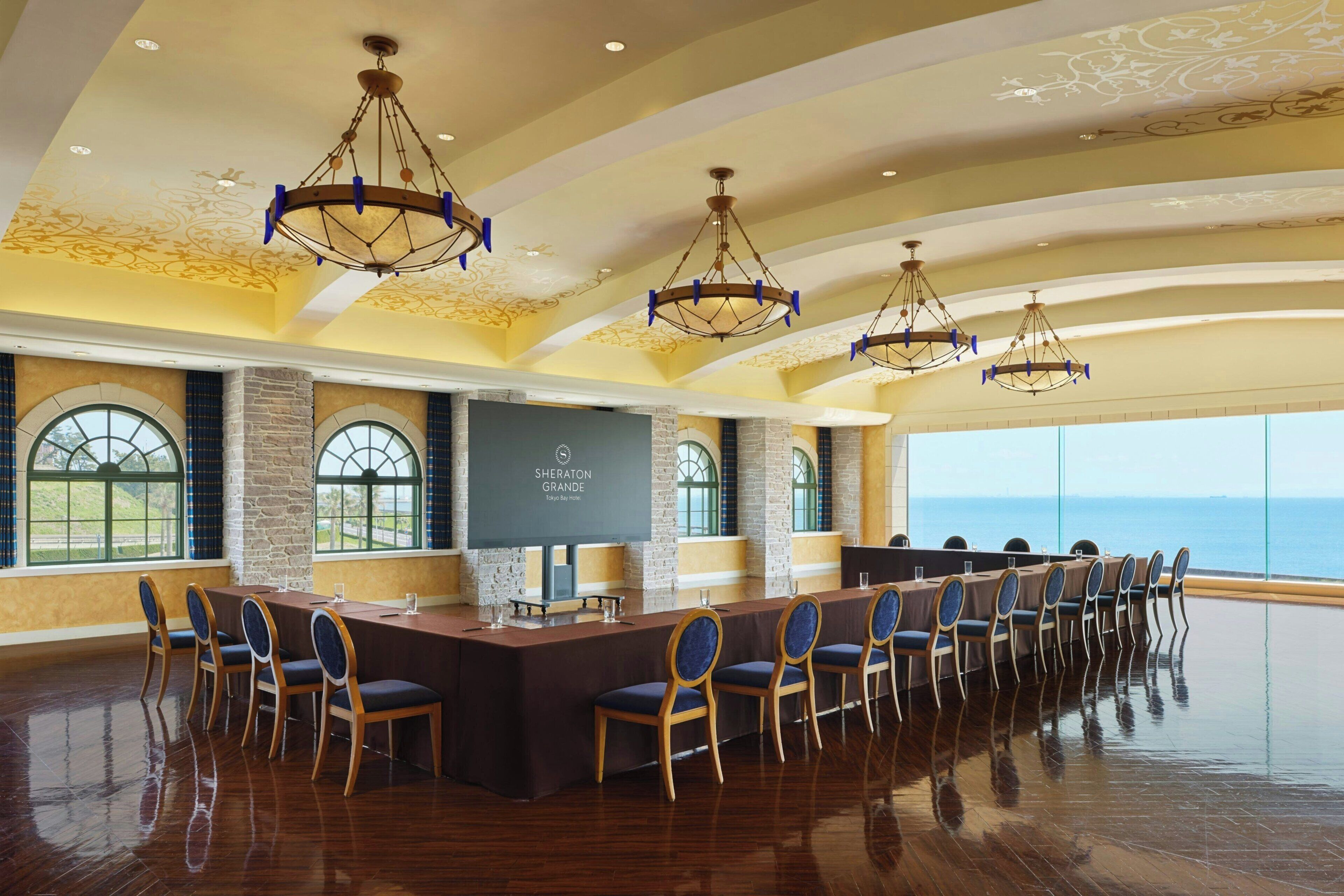 Uma sala de conferências com uma mesa comprida, cadeiras, lustres e vista para o oceano.