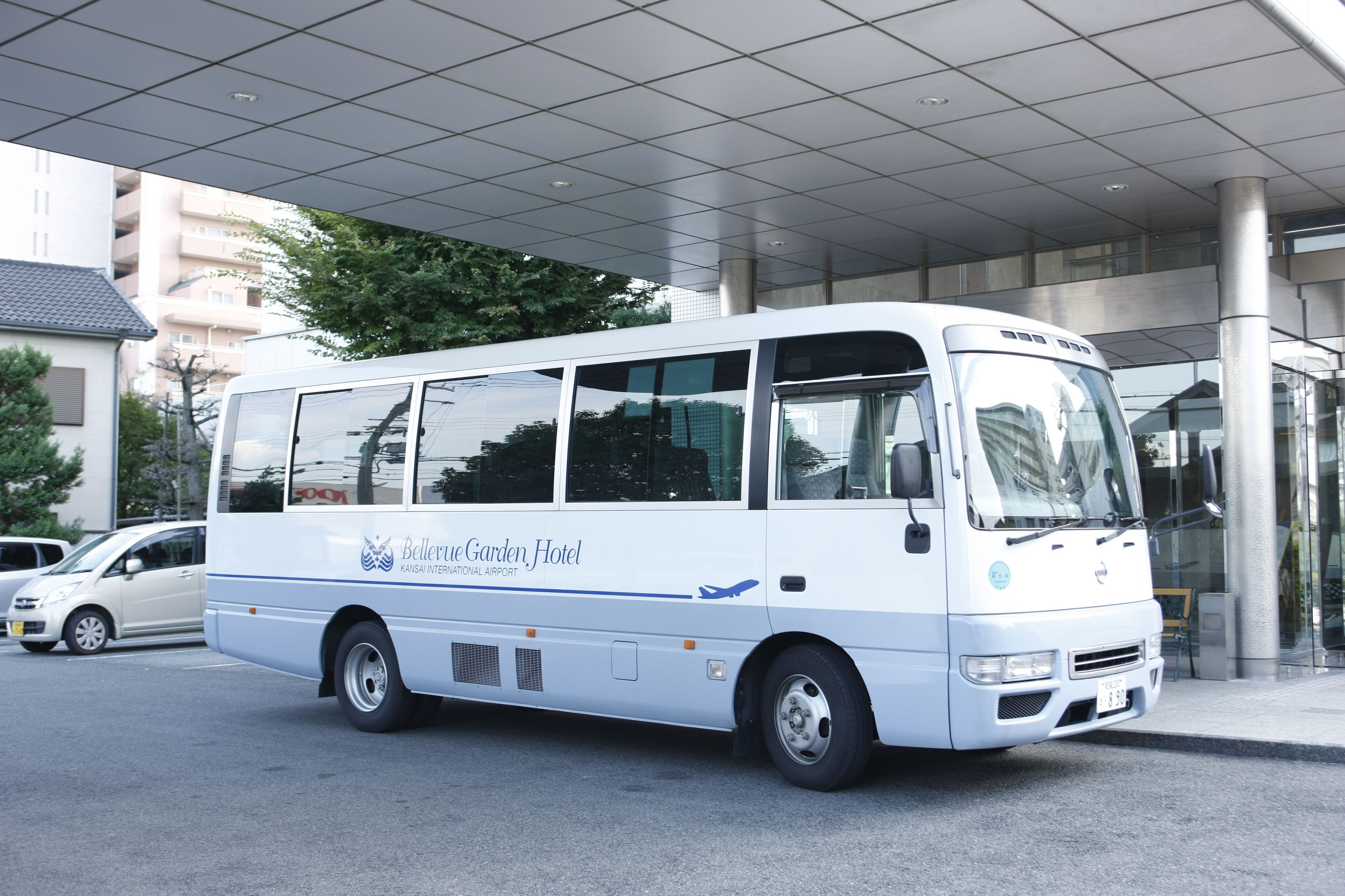 Um ônibus branco com a inscrição 'Bellflower Garden Hotel' e 'Aeroporto Internacional de Kansai' estacionado em frente a um edifício.