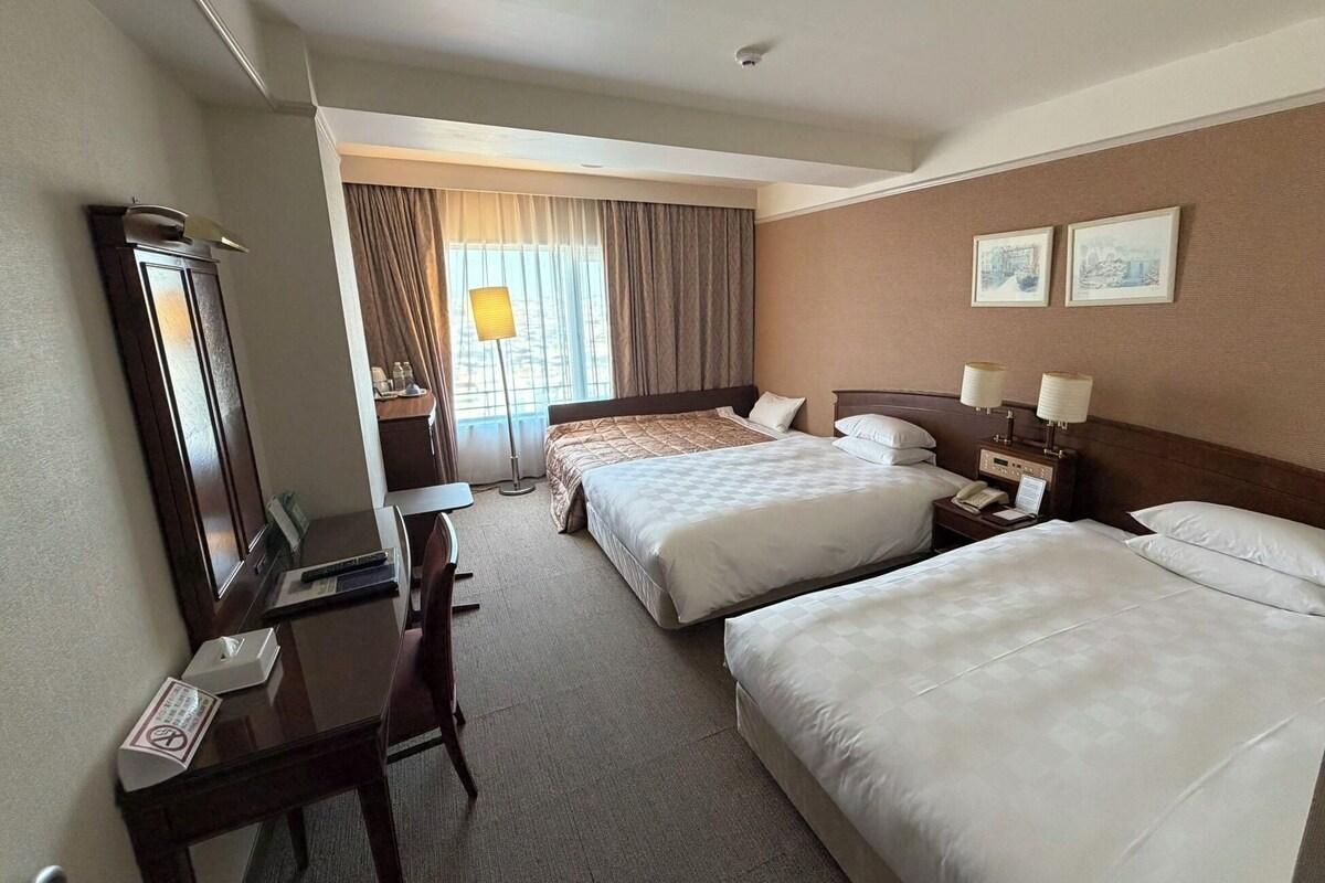 Quarto de hotel com duas camas, uma escrivaninha, uma cadeira, um abajur, um telefone e uma janela com cortinas.