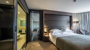 Quarto de hotel moderno com uma cama grande, mesa de cabeceira com abajur, uma cadeira e vista para o corredor e banheiro.